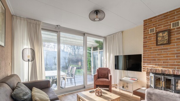 Uma sala de estar moderna com um sofá, poltrona e mesa de centro. Há uma lareira, uma televisão e uma janela grande com cortinas.