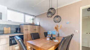 Uma cozinha moderna com armários de madeira, uma mesa de jantar e cadeiras pretas. Há luminárias pendentes, uma máquina de café e um vaso com plantas secas.