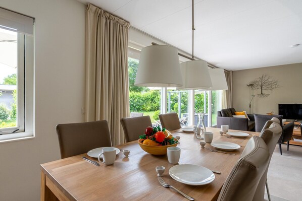 Uma área de refeições com uma mesa de madeira posta para quatro pessoas, uma tigela de frutas e vista para o exterior.
