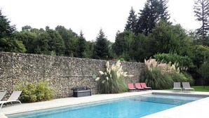 Piscina retangular com muro de pedra e plantas em vasos. Há duas espreguiçadeiras e um pequeno banco de madeira.