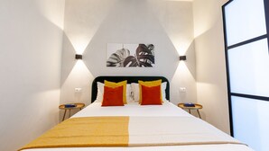 Cama bem arrumada com um cobertor amarelo, duas almofadas vermelhas e uma obra de arte em preto e branco acima. Há duas mesas de cabeceira com luminárias.