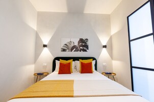 Cama bem arrumada com um cobertor amarelo, duas almofadas vermelhas e uma obra de arte em preto e branco acima. Há duas mesas de cabeceira com luminárias.