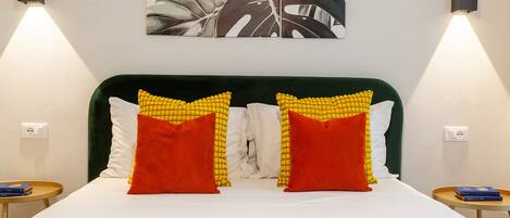 Uma cama com cabeceira estofada verde, duas almofadas padronadas em amarelo e vermelho, e um cobreleito branco.