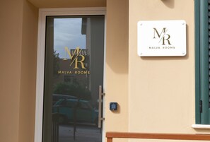 Entrada de hotel com porta de vidro exibindo o logotipo e o nome "Malva Rooms".