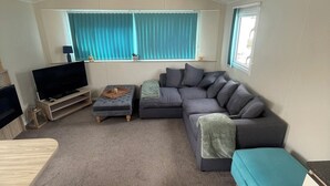 Uma sala de estar com um sofá seccional, uma TV em um móvel, um pequeno banquinho e uma janela com persianas.