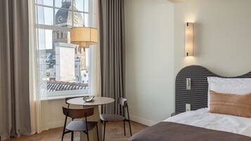 Quarto de hotel com cama, mesa redonda, duas cadeiras, janela com cortinas e vista para a cidade.