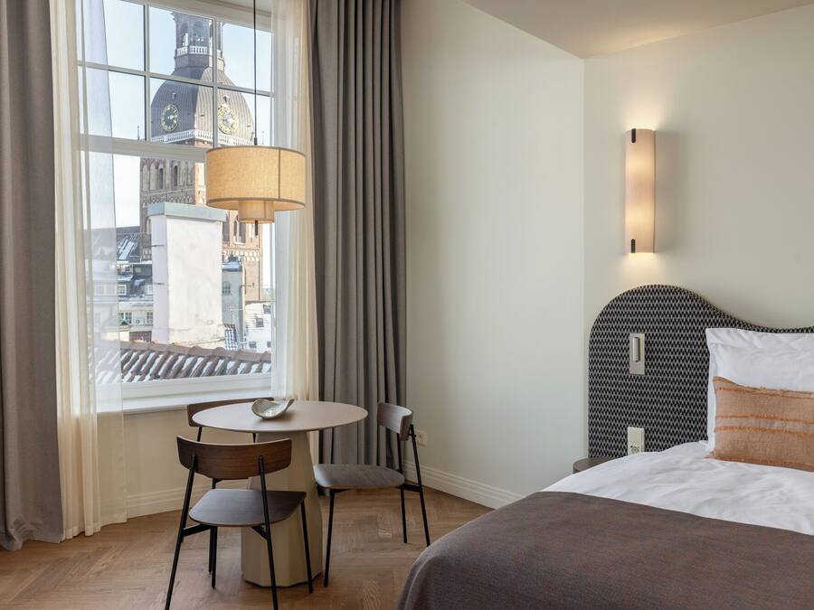 Quarto de hotel com cama, mesa redonda, duas cadeiras, janela com cortinas e vista para a cidade.