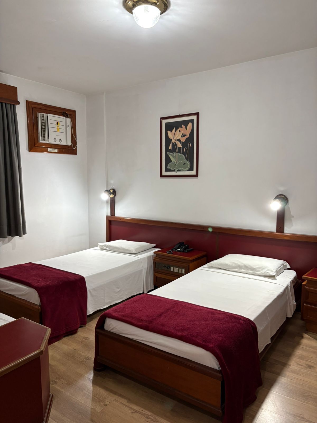 Quarto de hotel com duas camas de solteiro, cabeceira de madeira, mesa de cabeceira com abajur, ilustração botânica emoldurada e tomada na parede.