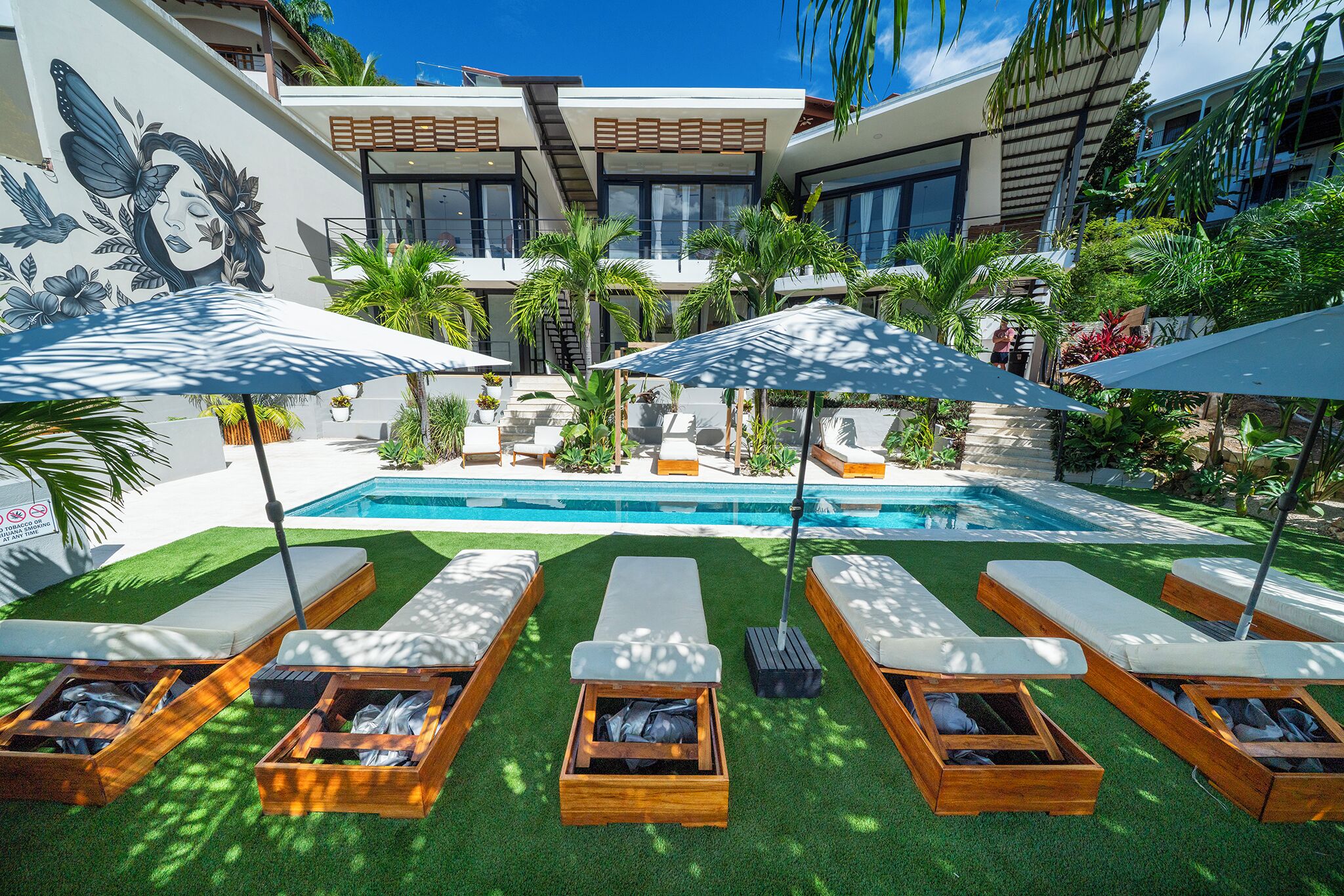 Uma área moderna de piscina ao ar livre com espreguiçadeiras e guarda-sóis, cercada por vegetação exuberante e um mural decorativo.