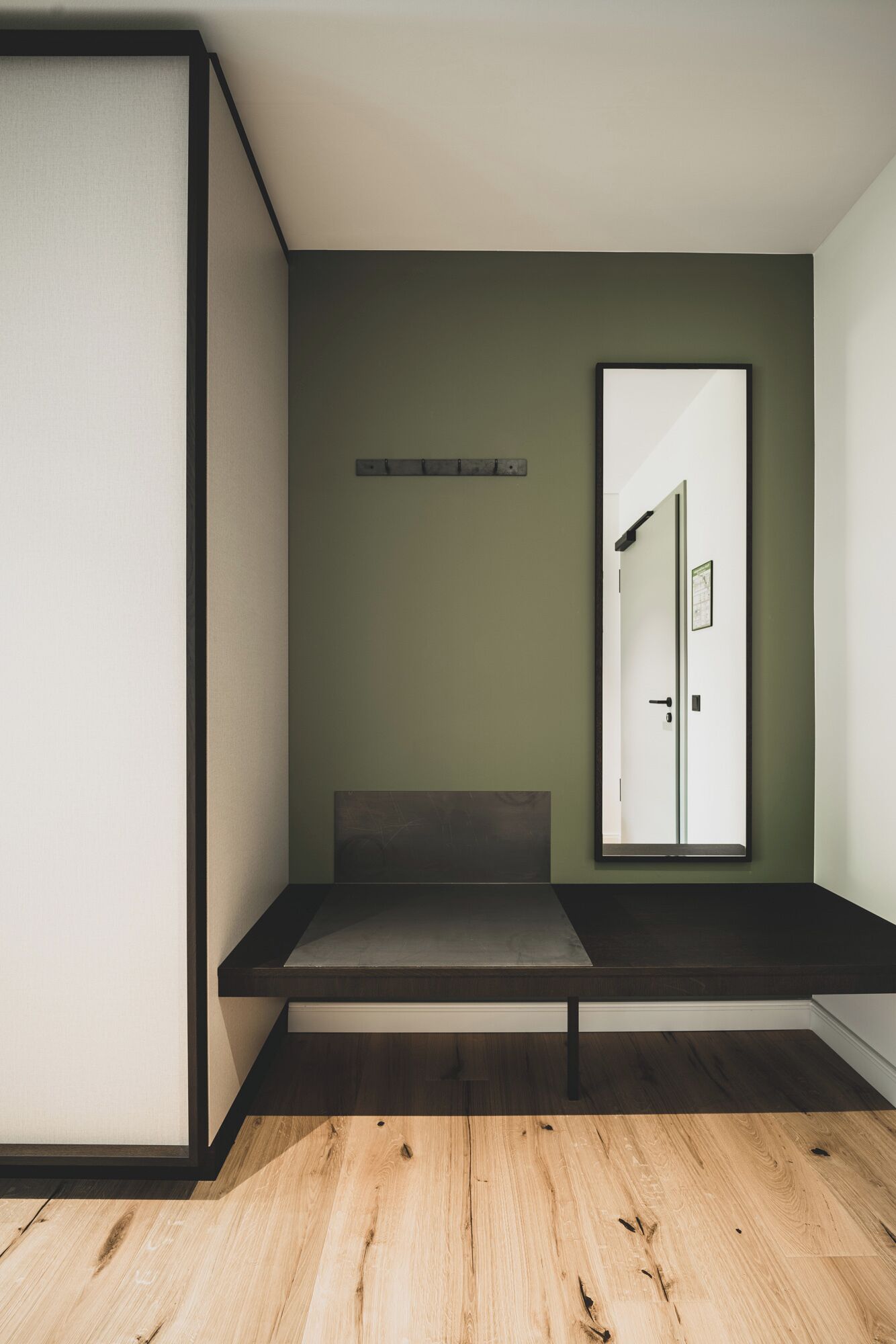 Quarto minimalista com piso de madeira, um banco, parede verde e porta com espelho.