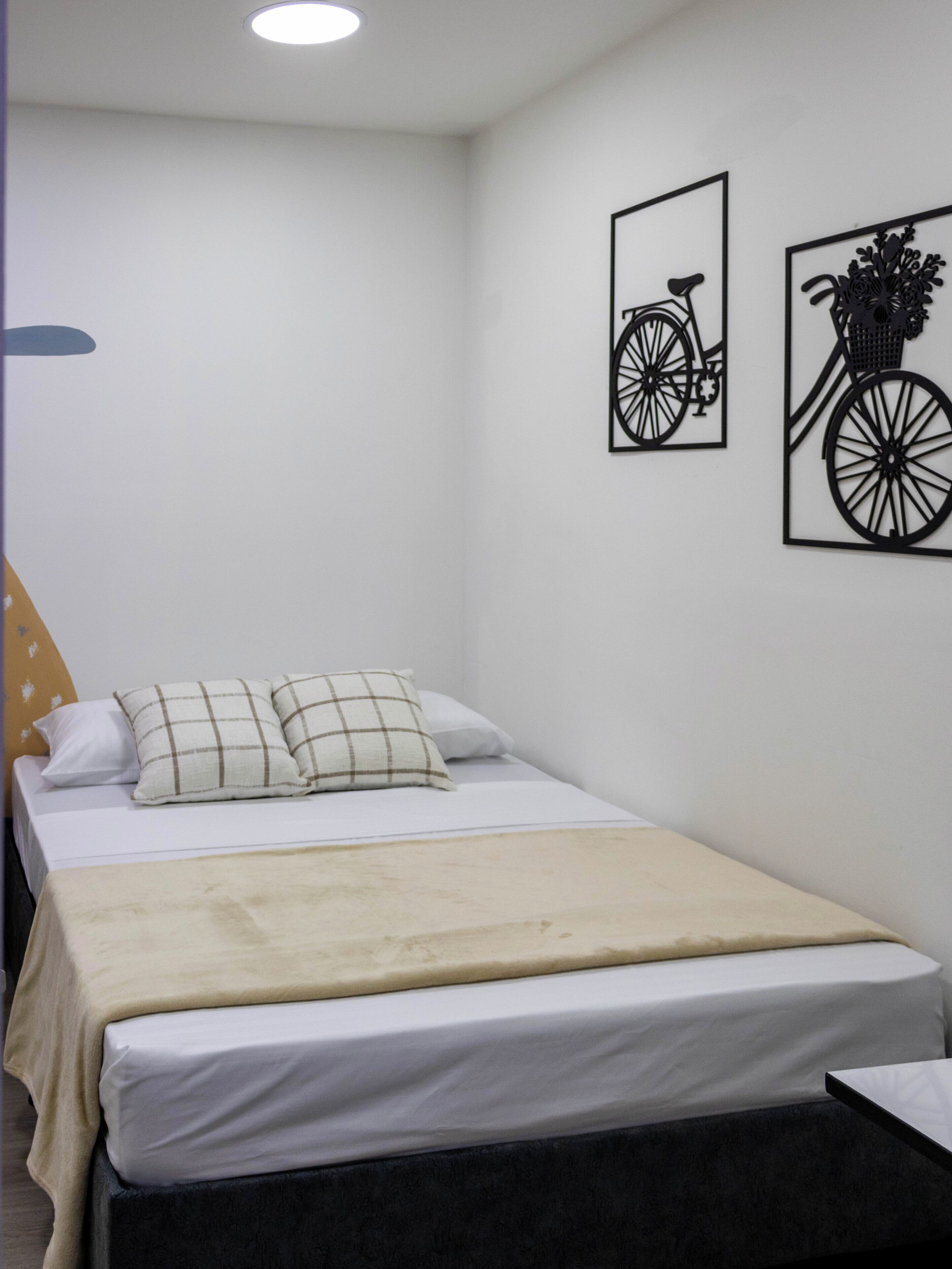 Cama bem arrumada com edredom branco e bege, cabeceira preta e um único travesseiro. Duas obras de arte emolduradas nas paredes retratam bicicletas.