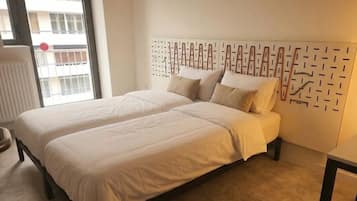 Uma cama de casal com cabeceira com padrão geométrico, janela com vista para um prédio e um radiador à esquerda.