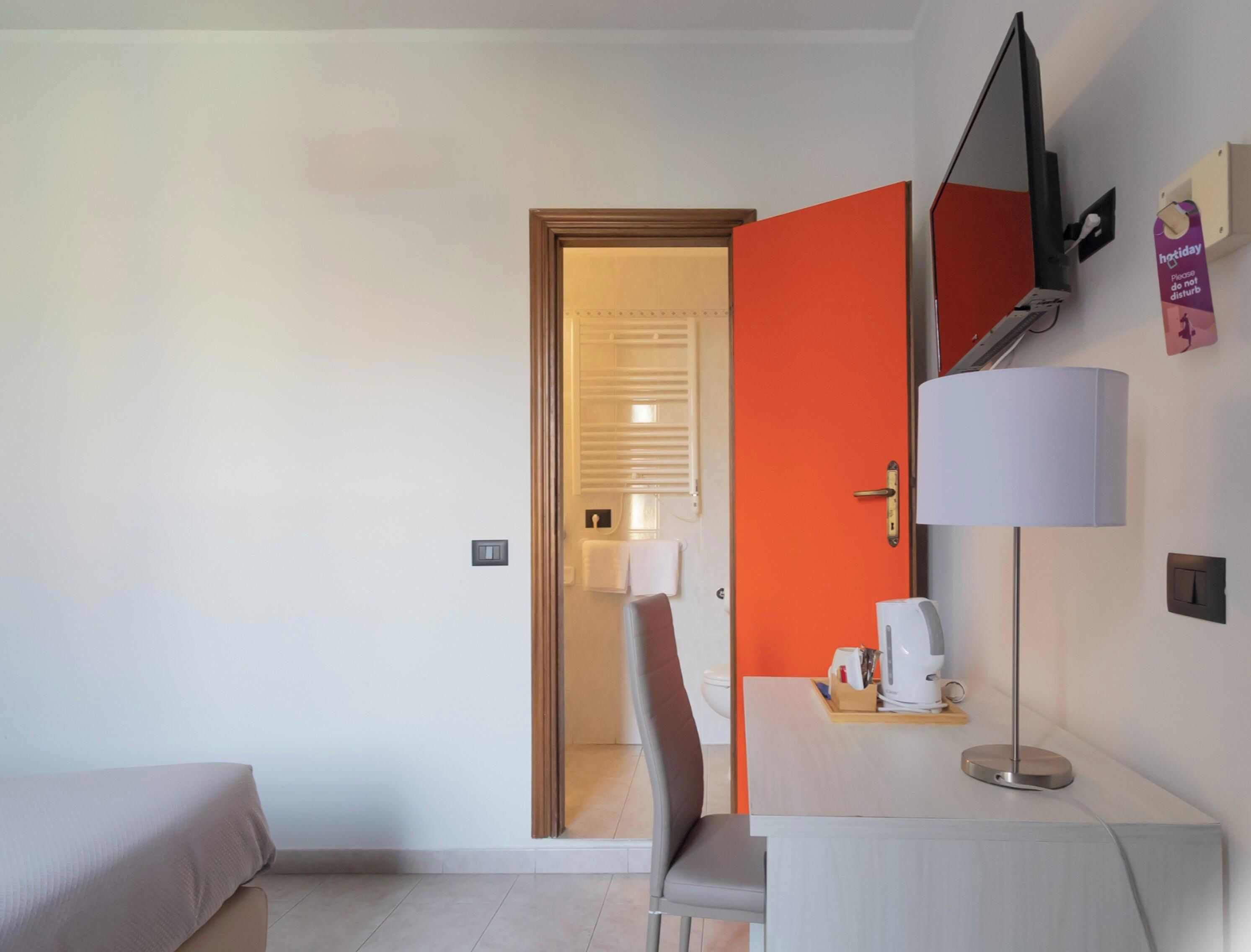 Quarto de hotel com cama, escrivaninha com luminária, televisão e banheiro visível por uma porta aberta.