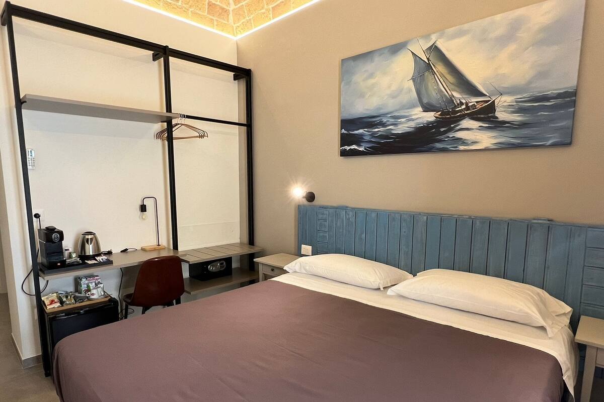 Quarto de hotel com cama, escrivaninha, cadeira, máquina de café e quadro de um veleiro na parede.