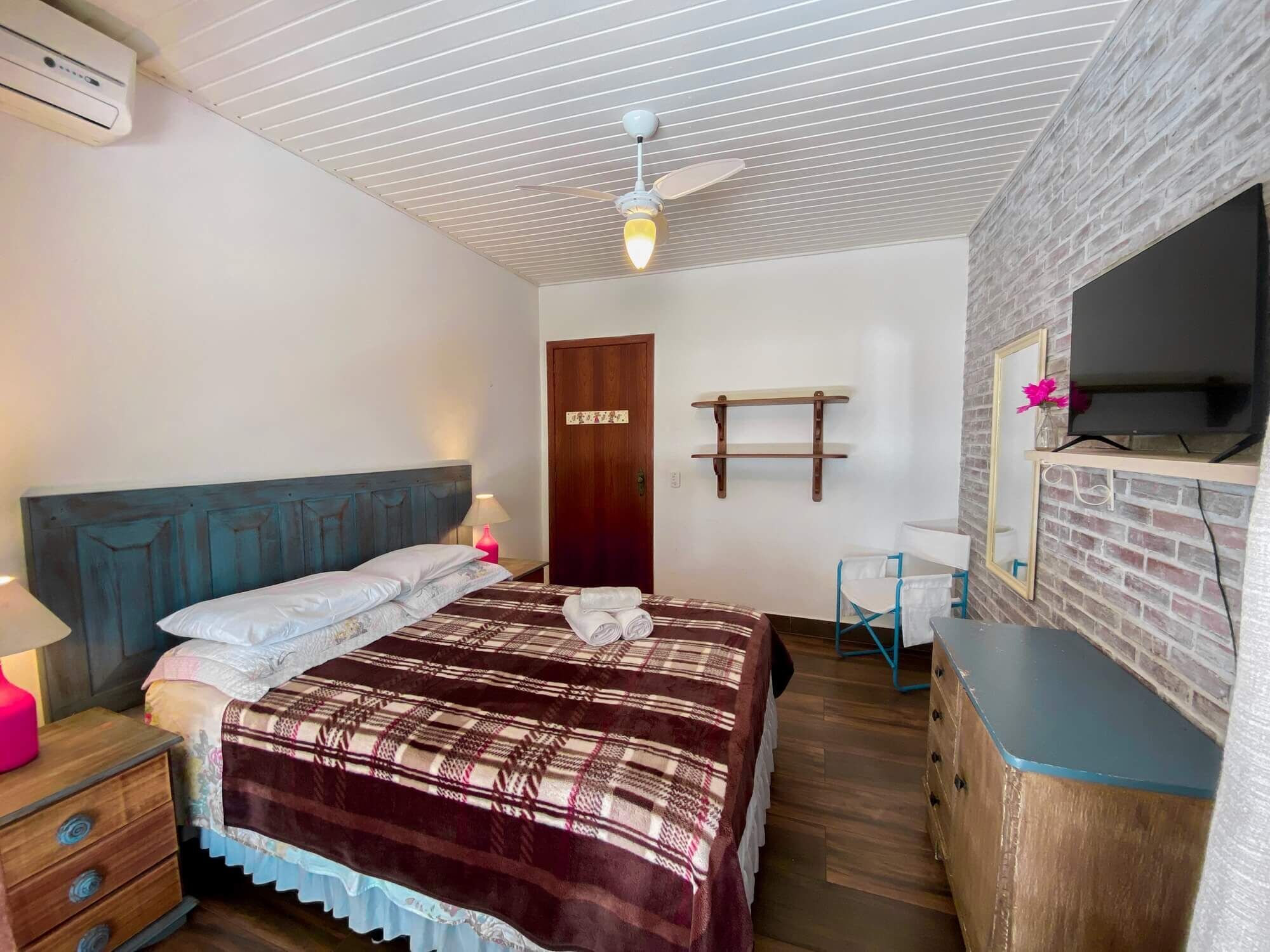 Quarto com cama, mesa de cabeceira, abajur, guarda-roupa e televisão fixada numa parede de tijolos.