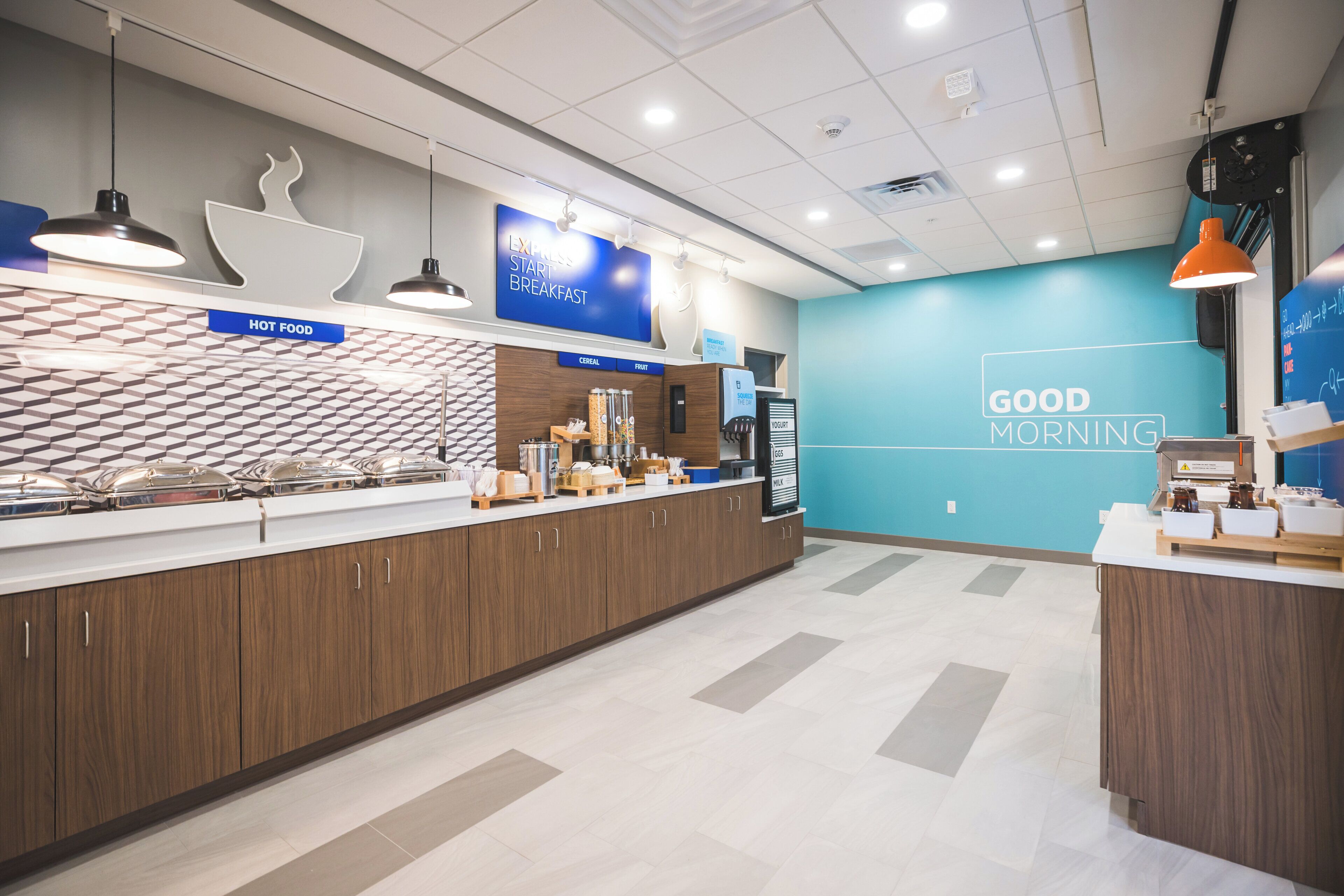 Espaço de café da manhã com balcão, distribuidores de alimentos e parede com a placa 'GOOD MORNING'.