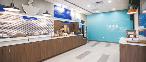 Espaço de café da manhã com balcão, distribuidores de alimentos e parede com a placa 'GOOD MORNING'.