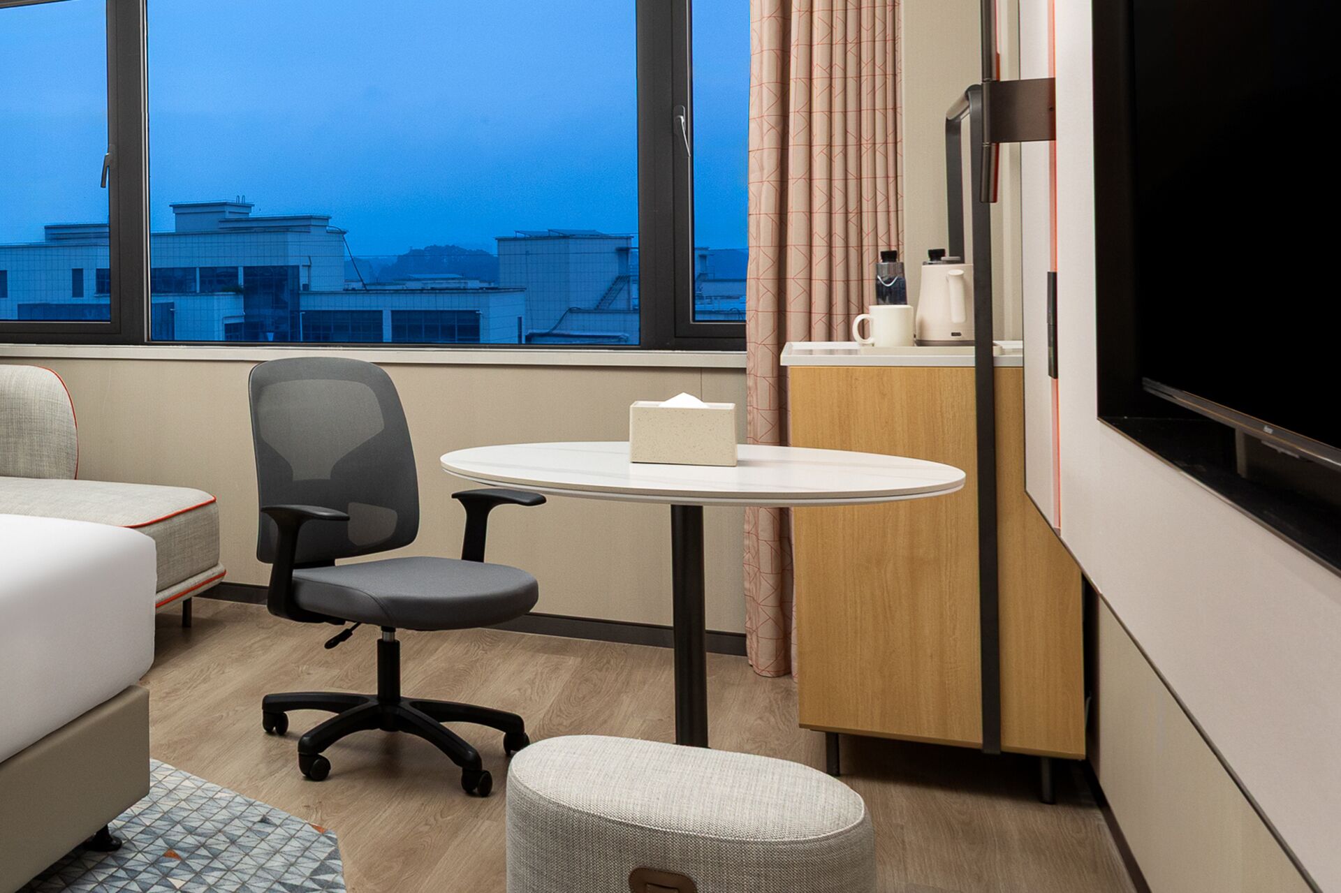 Quarto de hotel com uma cama, uma escrivaninha, uma cadeira, uma mesa redonda pequena, uma televisão e uma janela com vista para a cidade.