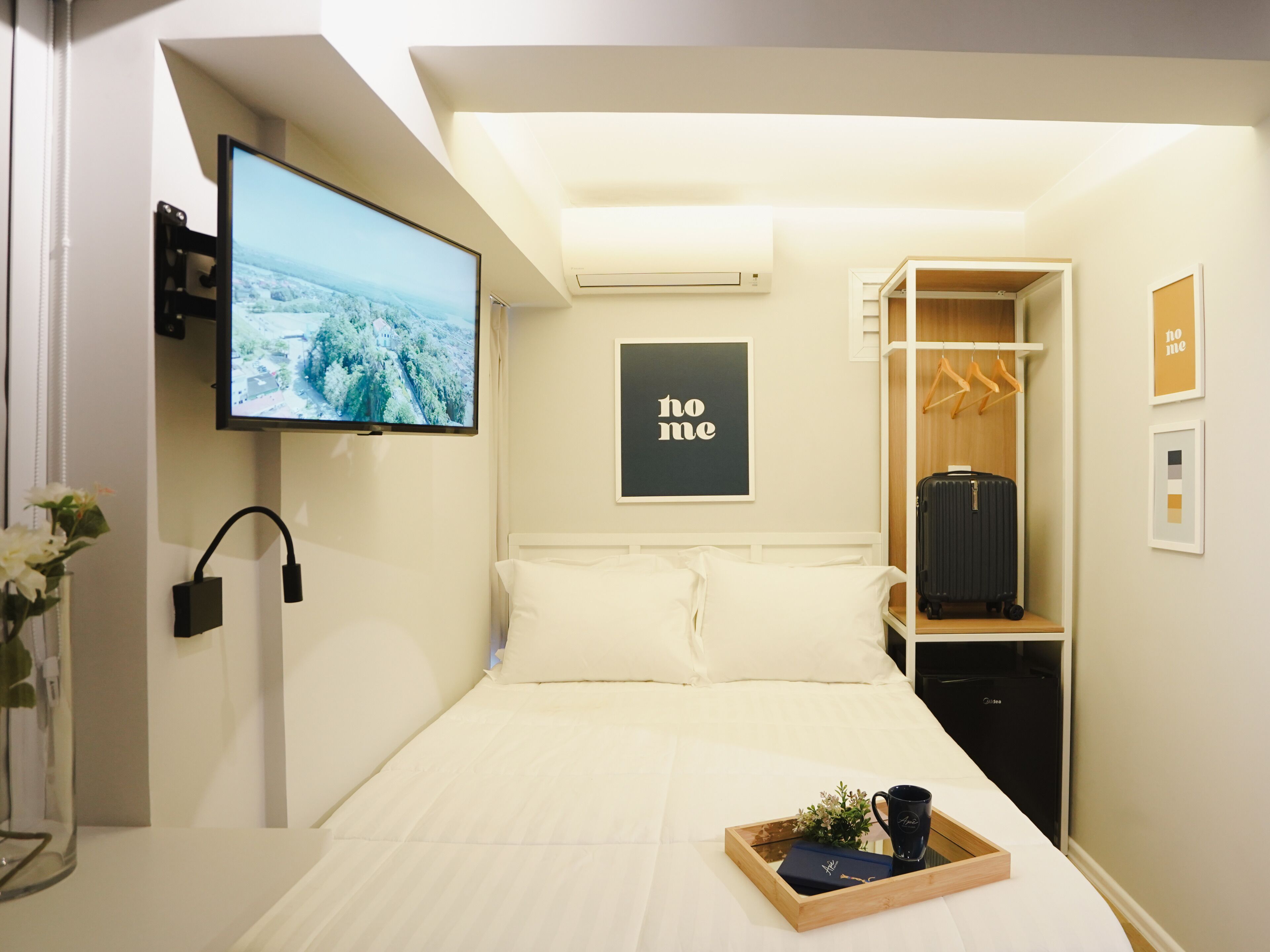 Quarto de hotel com cama, televisão fixada na parede, guarda-roupa e um letreiro emoldurado com a palavra 'home'.
