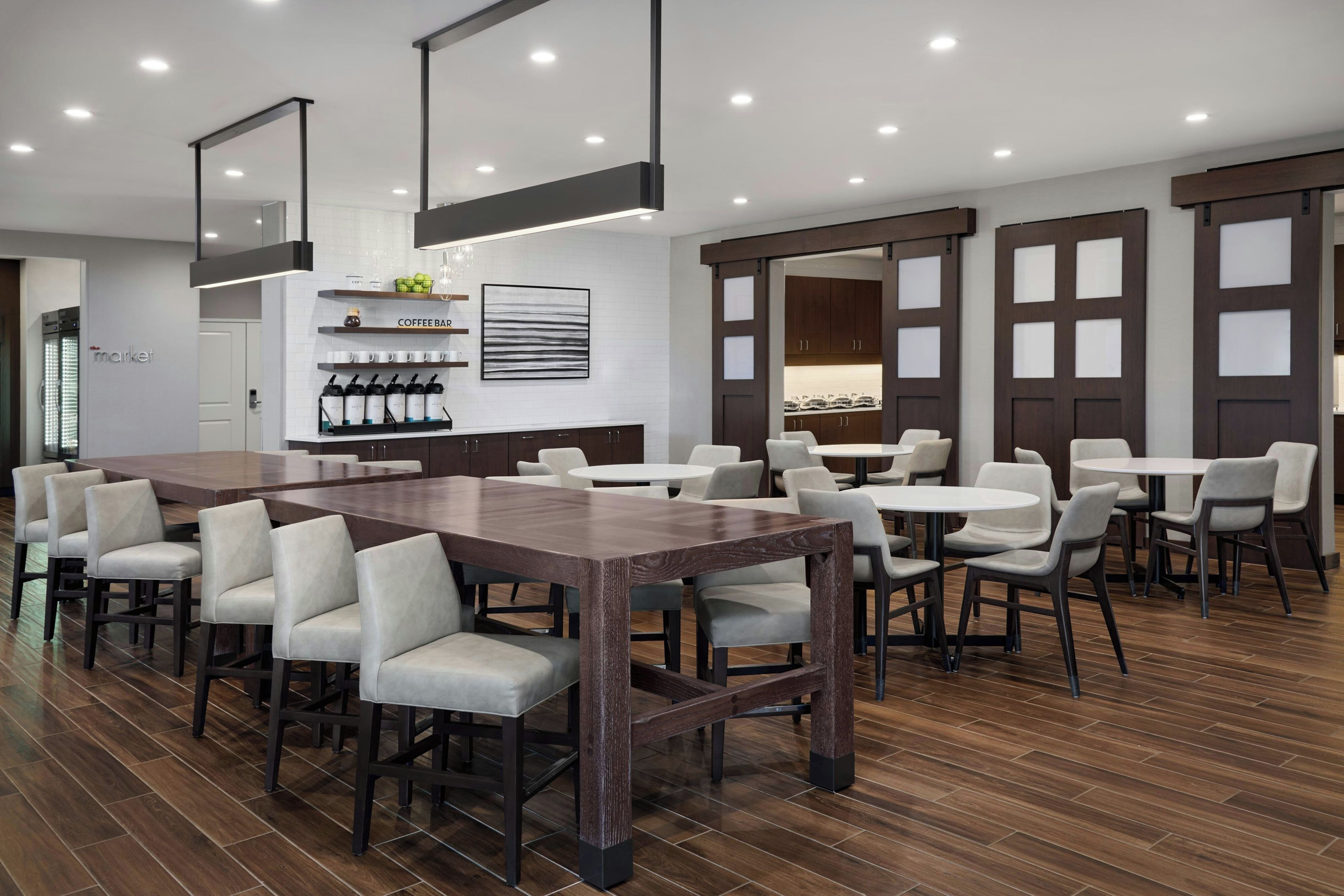 Uma sala de jantar moderna com uma longa mesa de madeira e várias cadeiras, um bar de café com diversos equipamentos para preparo de café, e uma cozinha com armários e geladeira.