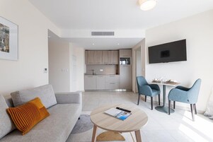 Uma sala de estar moderna com um sofá cinza, uma mesa de centro redonda e uma área de jantar com duas cadeiras azuis e uma mesinha.