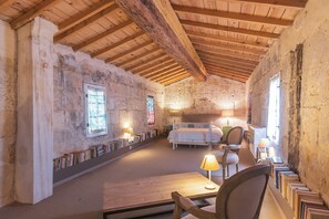 Quarto rústico com vigas de madeira aparente, uma cama, uma escrivaninha com abajur e estantes.