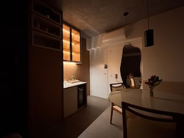 Uma área de refeições compacta com uma mesa redonda, cadeiras, uma cozinha pequena com prateleiras iluminadas e uma porta branca.