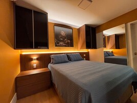 Quarto com cama, mesas de cabeceira, televisão presa à parede, quadro emoldurado e espelho.