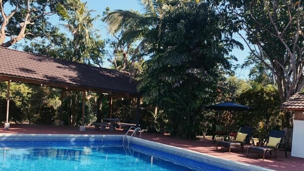 Piscina com borda azulejada, rodeada por espreguiçadeiras e uma área de estar coberta.