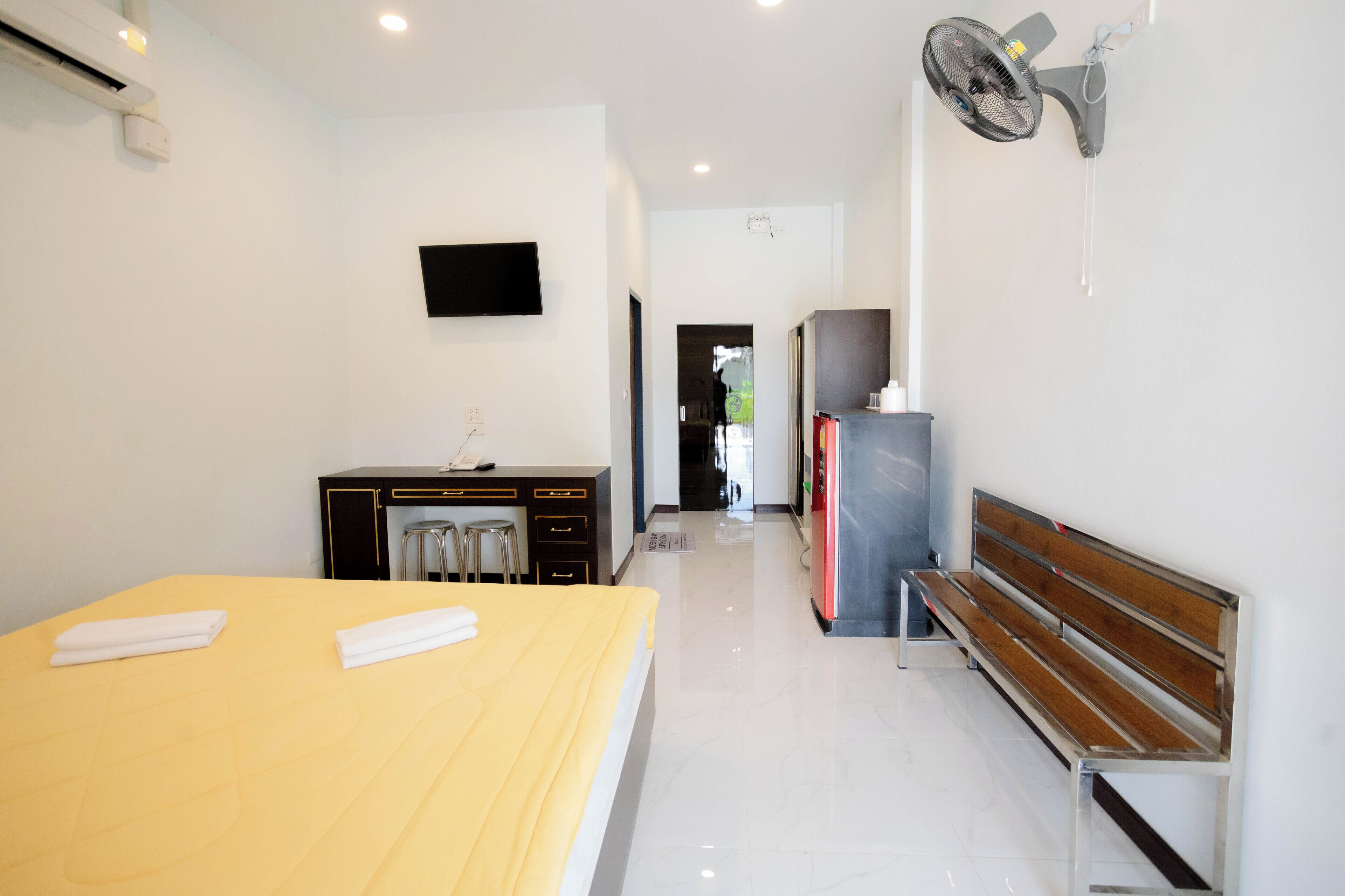 Quarto de hotel com uma cama, uma escrivaninha com cadeira, um banco, um frigorífico e um micro-ondas, além de um ventoinha fixado na parede.