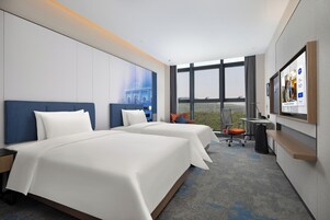 Quarto de hotel com duas camas, uma escrivaninha, uma cadeira e uma janela grande com vista.