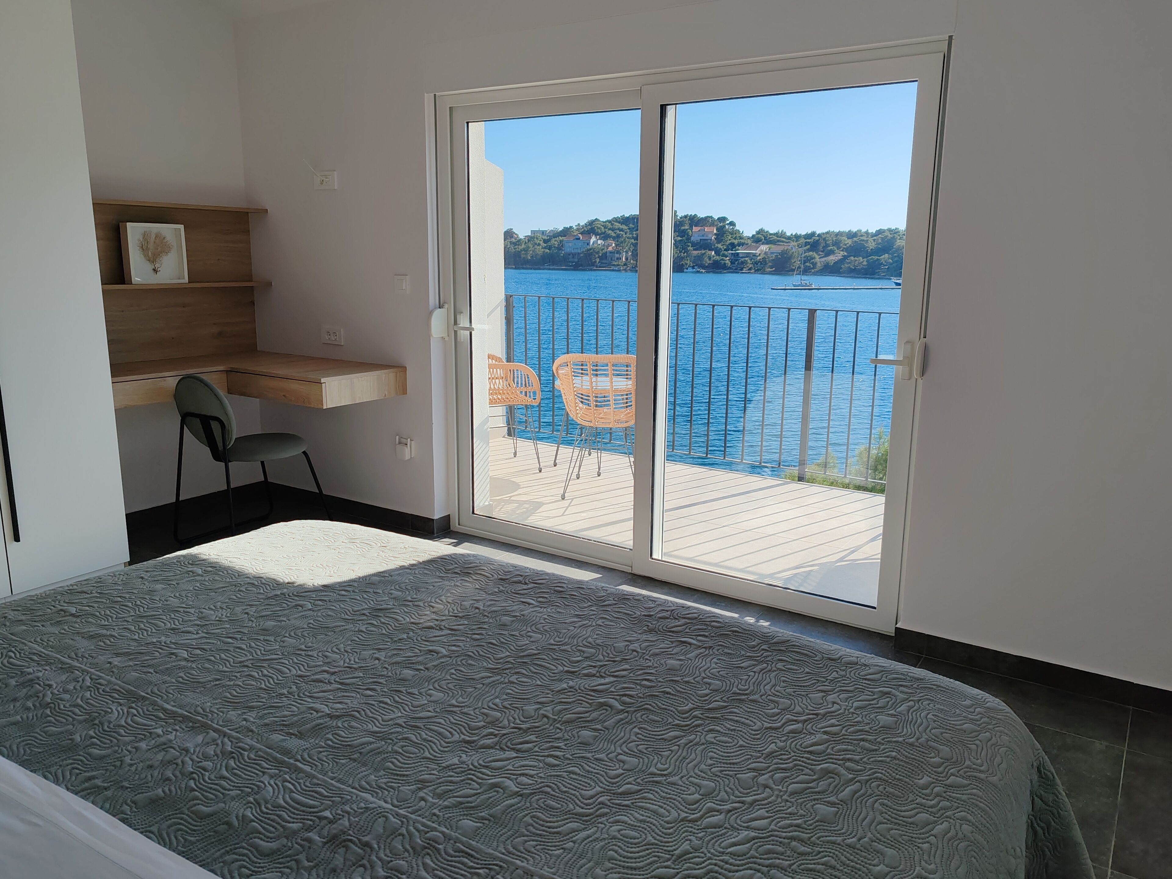 Quarto com cama, escrivaninha e cadeira. Porta de correr dá acesso a uma varanda com vista para um corpo de água e edifícios.