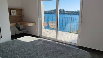 Quarto com cama, escrivaninha e cadeira. Porta de correr dá acesso a uma varanda com vista para um corpo de água e edifícios.