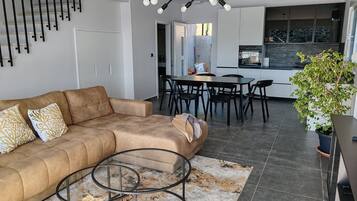 Uma sala de estar moderna com um sofá bege, uma mesa de centro de vidro e uma área de jantar com mesa e cadeiras pretas. É possível ver uma cozinha ao fundo, com uma janela grande.