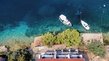 Vista aérea de uma área costeira com duas embarcações ancoradas em águas azuis cristalinas, uma costa rochosa com árvores e um edifício com telhado vermelho.