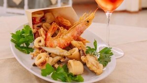 Um prato de mariscos com um copo de vinho tinto sobre uma mesa em um restaurante com decoração clara e portas arqueadas.