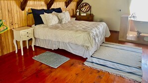 Quarto com teto de madeira, cama com roupa de cama branca, mesa de cabeceira, cômoda, espelho e banheiro com banheira.