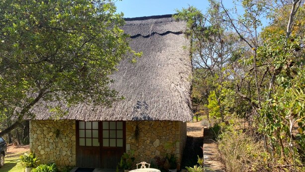 Uma casa de pedra com telhado de palha, rodeada por árvores e um pequeno jardim com mesa e cadeiras.
