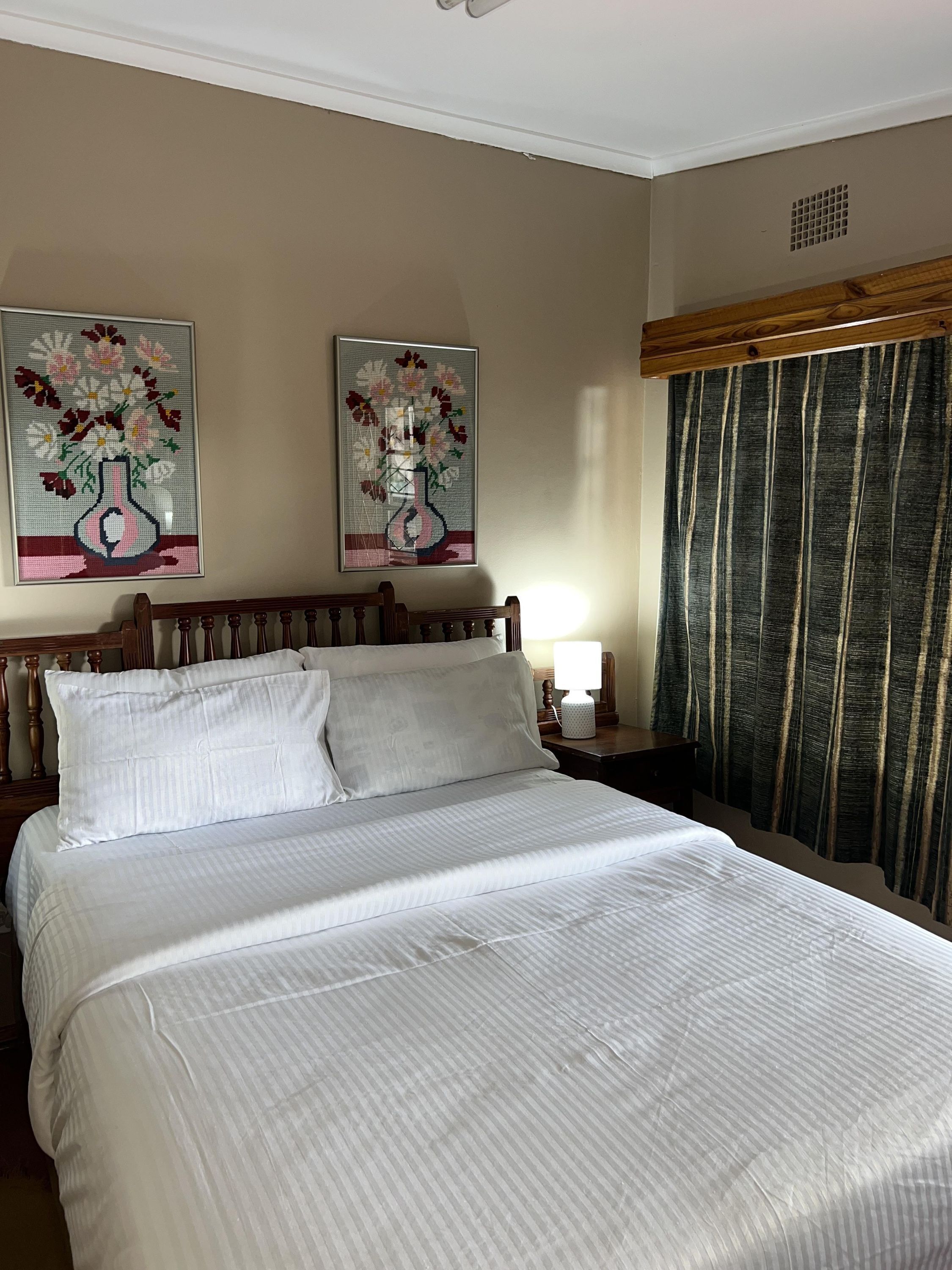 Quarto de hotel com uma cama grande, duas pinturas penduradas na parede, uma cabeceira de madeira, uma mesa de cabeceira com uma luminária e uma janela com cortinas listradas.