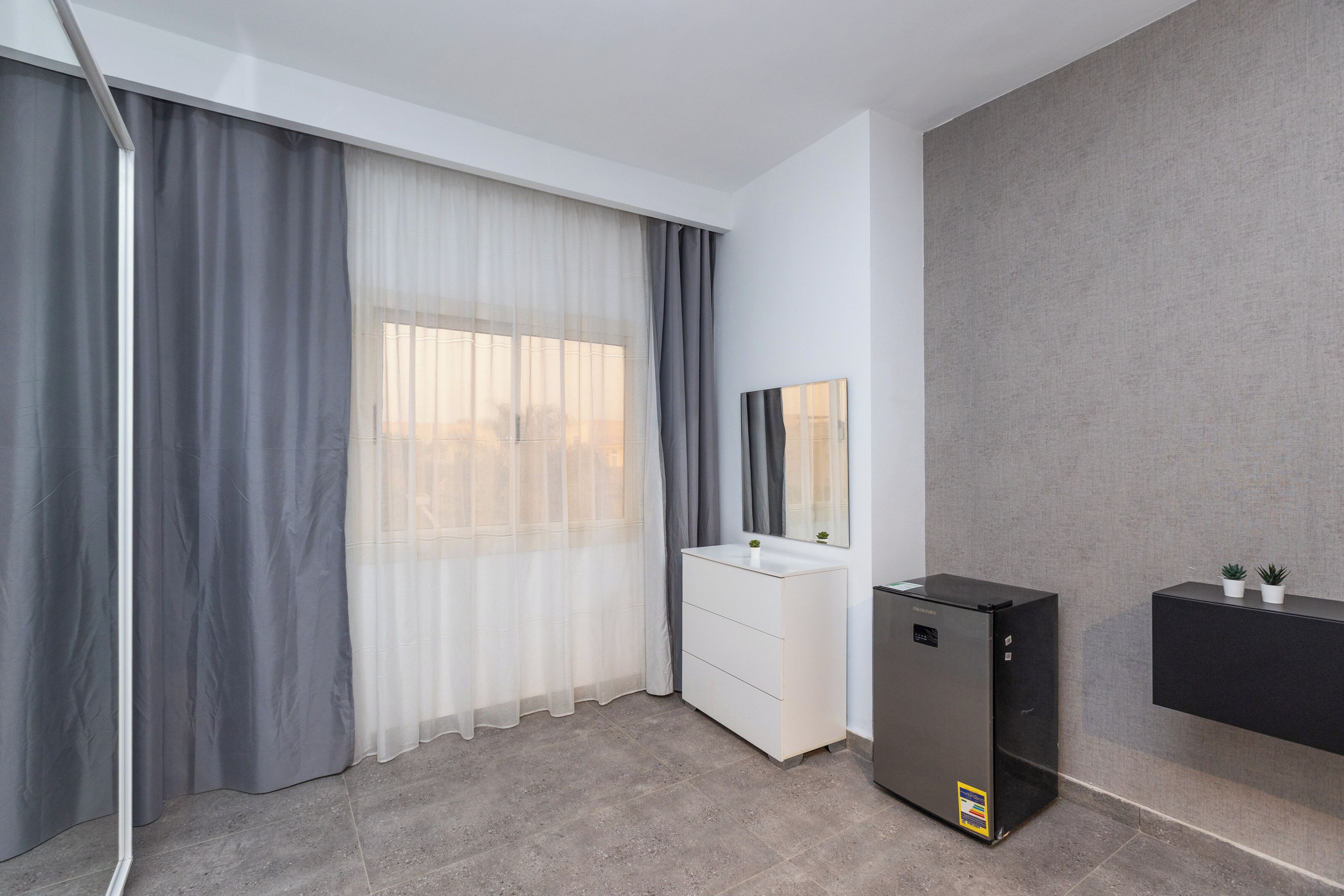 Quarto de hotel moderno com uma janela grande, penteadeira branca, minibar preto e parede cinza de destaque.