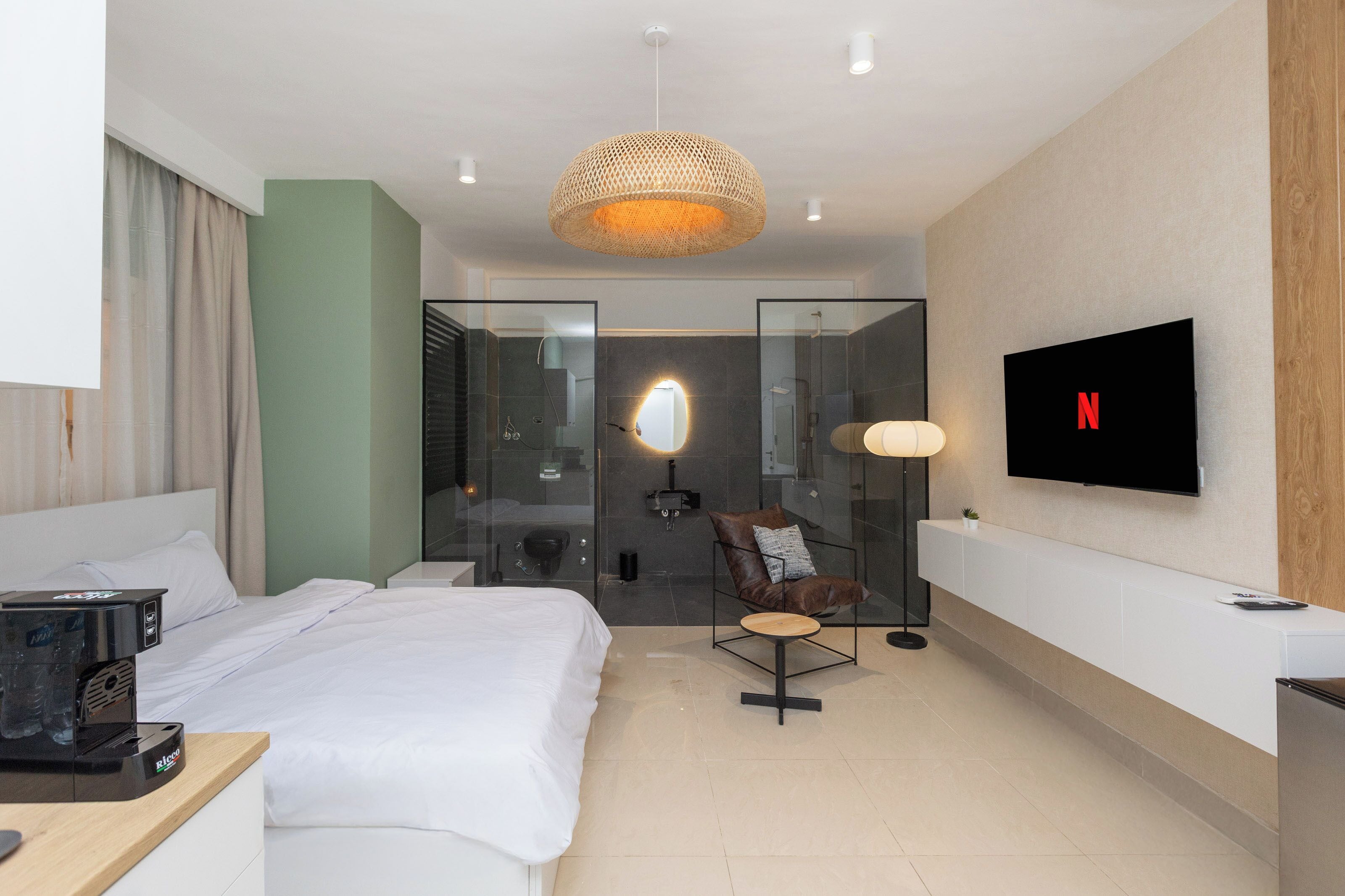 Quarto de hotel moderno com cama, cafeteira, área de duche com paredes de vidro, poltrona de couro, TV de ecrã plano e logótipo da Netflix fixado na parede.