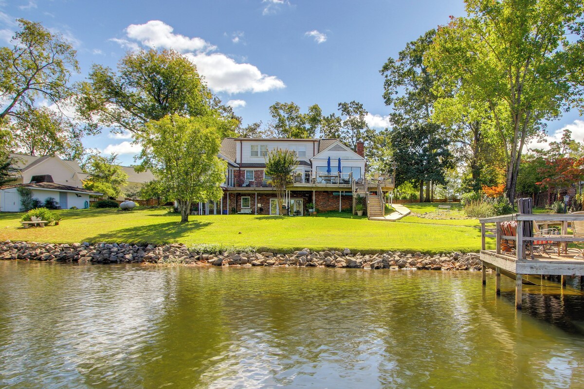Uma casa com um píer que se estende por um lago calmo, rodeada por vegetação exuberante e um céu limpo.
