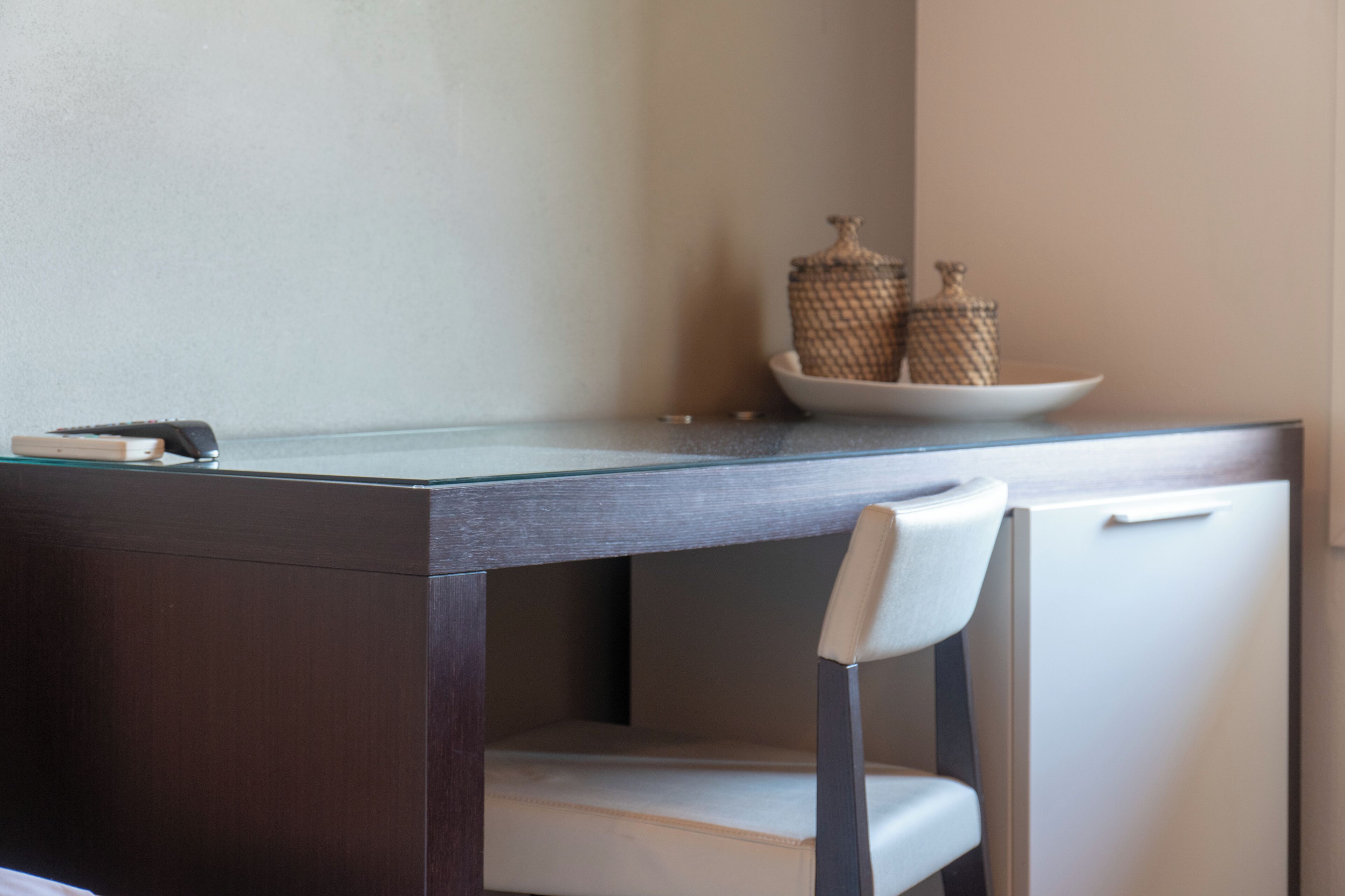 Uma secretária moderna com tampo de vidro, uma cadeira branca e objetos decorativos ao lado.