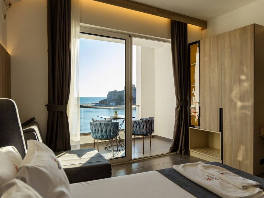 Quarto de hotel com cama, uma mesinha, uma cadeira e vista para o mar.