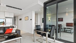 Uma sala de estar moderna com uma mesa de jantar de vidro, sofá de couro preto e uma porta de correr de vidro que dá para uma varanda.