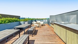 Terraço no telhado com bancos de madeira, uma mesa, cadeiras, área de bar e vista do horizonte urbano.