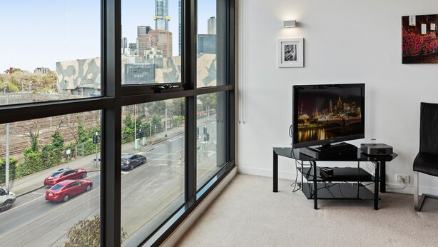 Quarto moderno com uma grande janela com vista para a paisagem urbana, uma televisão de tela plana e um espaço de trabalho minimalista.