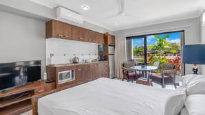 Quarto de hotel com cama grande, televisão, cozinha compacta e varanda com vista para áreas verdes e edifícios.