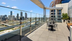 Terraço no telhado com guarda-corrimão de vidro, mesa com cadeiras e vista para o horizonte da cidade e um rio.