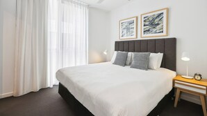 Quarto de hotel com uma cama grande, um ventilador de teto, duas obras de arte emolduradas, uma mesa de cabeceira com abajur e uma mesinha de noite com relógio.